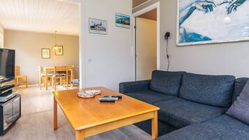 Uma sala de estar com uma mesa de madeira, um sofá cinzento, uma televisão e quadros nas paredes.