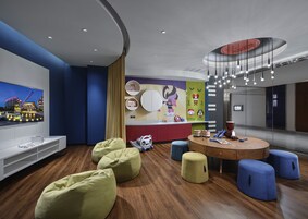 Uma sala de jogos infantil moderna, com uma televisão grande, puffs, uma mesa redonda e decorações coloridas nas paredes.