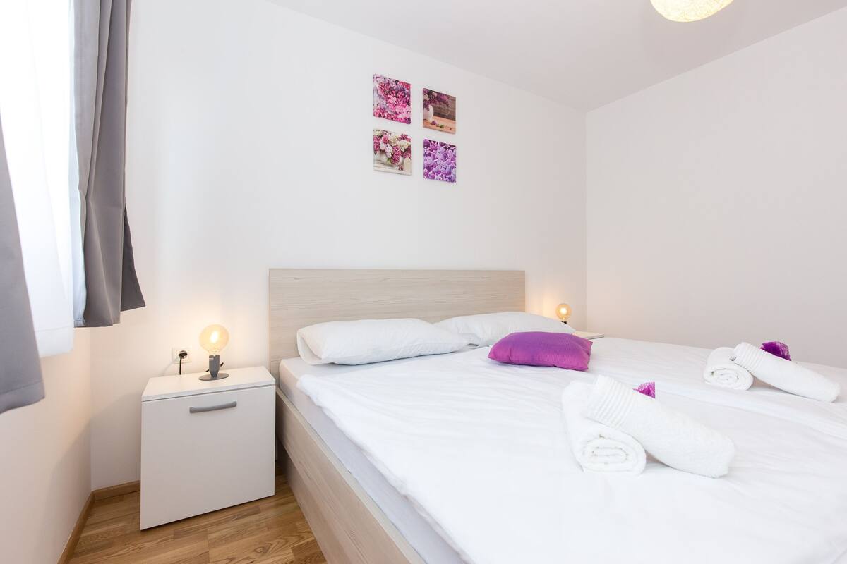 Cama bem arrumada com lençóis brancos e um travesseiro roxo, uma pequena mesa de cabeceira com abajur e uma cômoda com luminária.