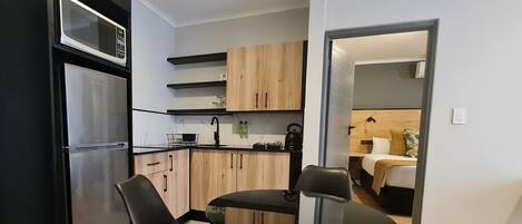 Cozinha compacta com geladeira, micro-ondas e armários de madeira. Há uma pequena mesa de jantar com duas cadeiras. Um quarto com cama e mesa de cabeceira é visível por uma porta aberta.