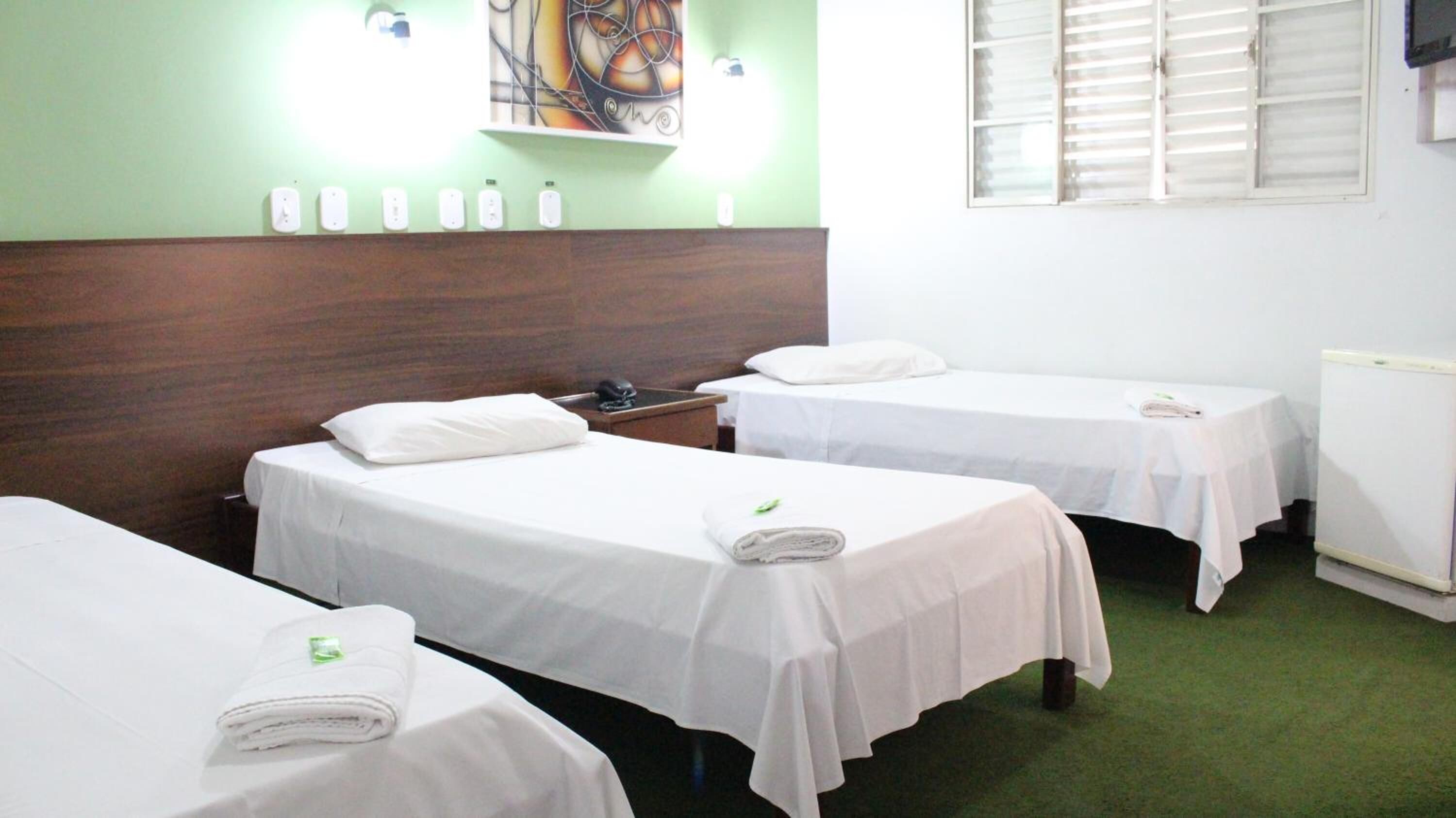 Quarto de hotel com duas camas de solteiro, cabeceira de madeira, mesa de cabeceira com abajur e uma obra de arte pendurada na parede.