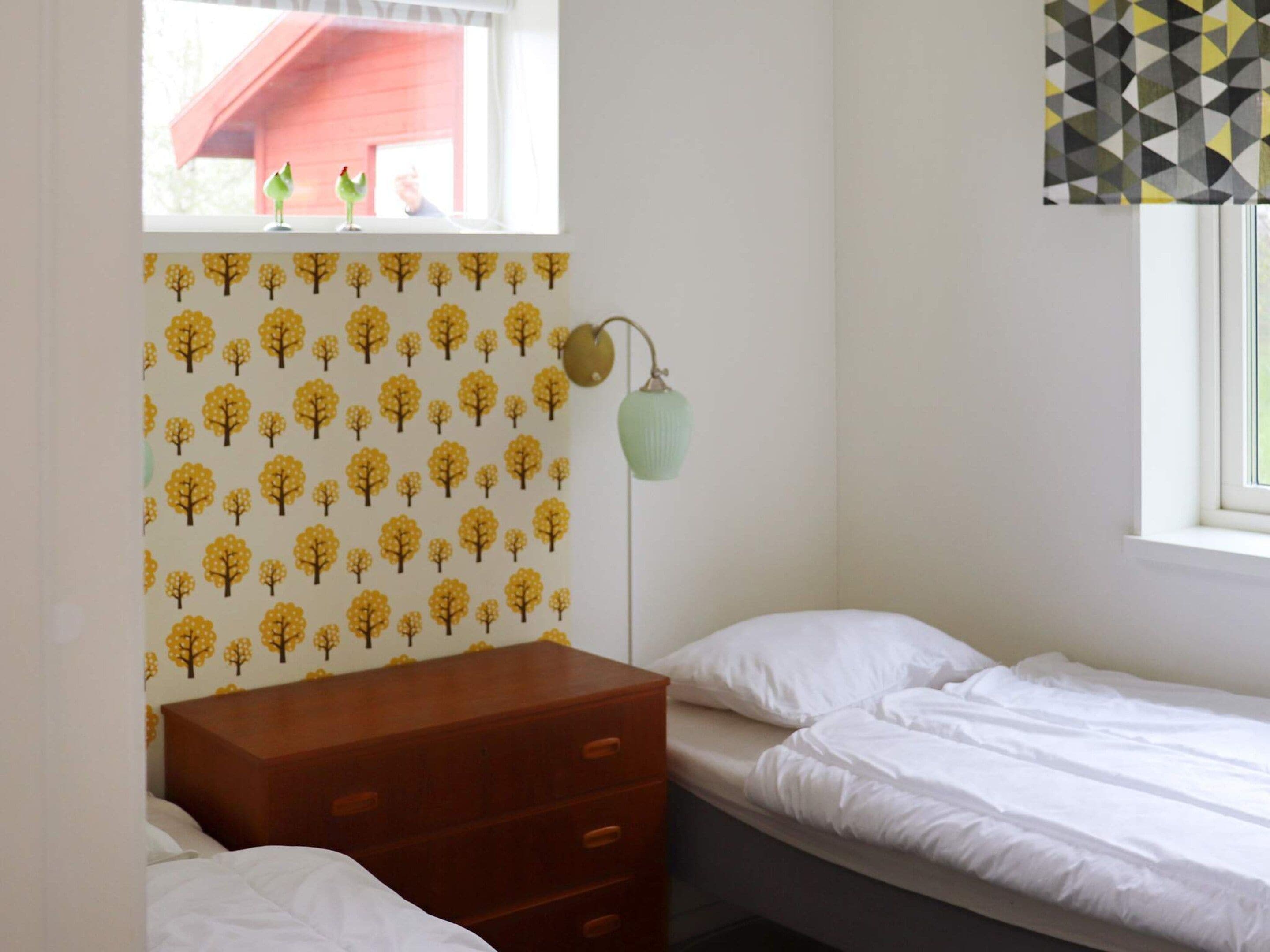 Quarto com uma cama de solteiro, cômoda, luminária na parede e janela com vista para um prédio vermelho.