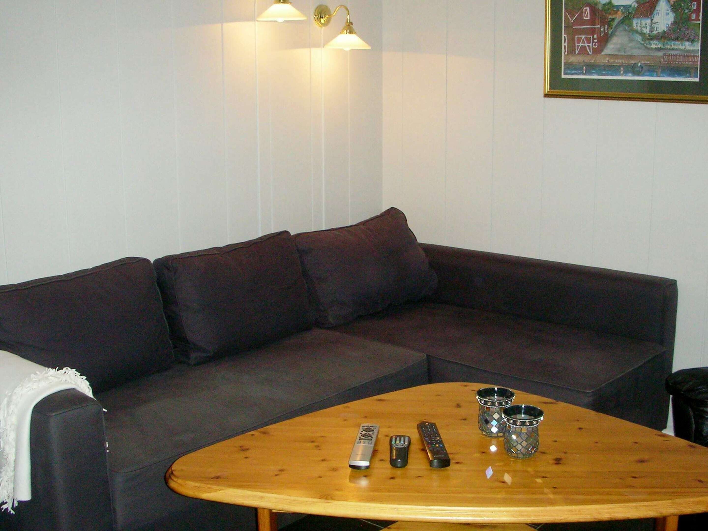 Uma sala de estar com um sofá marrom escuro, uma mesa de centro de madeira com controles remotos e objetos decorativos, e iluminação embutida na parede.