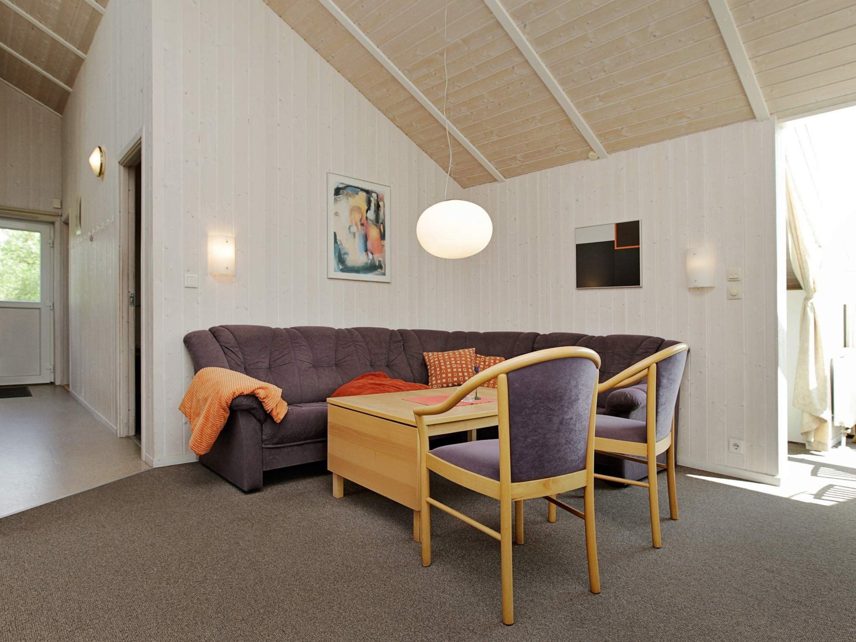 Uma sala de estar moderna com um sofá cinza escuro, uma mesa de centro de madeira e duas poltronas. Há um quadro na parede e uma luminária pendente sobre a mesa.