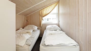 Uma cabana pequena, com um único quarto, duas camas, paredes de madeira e uma janela com cortinas.
