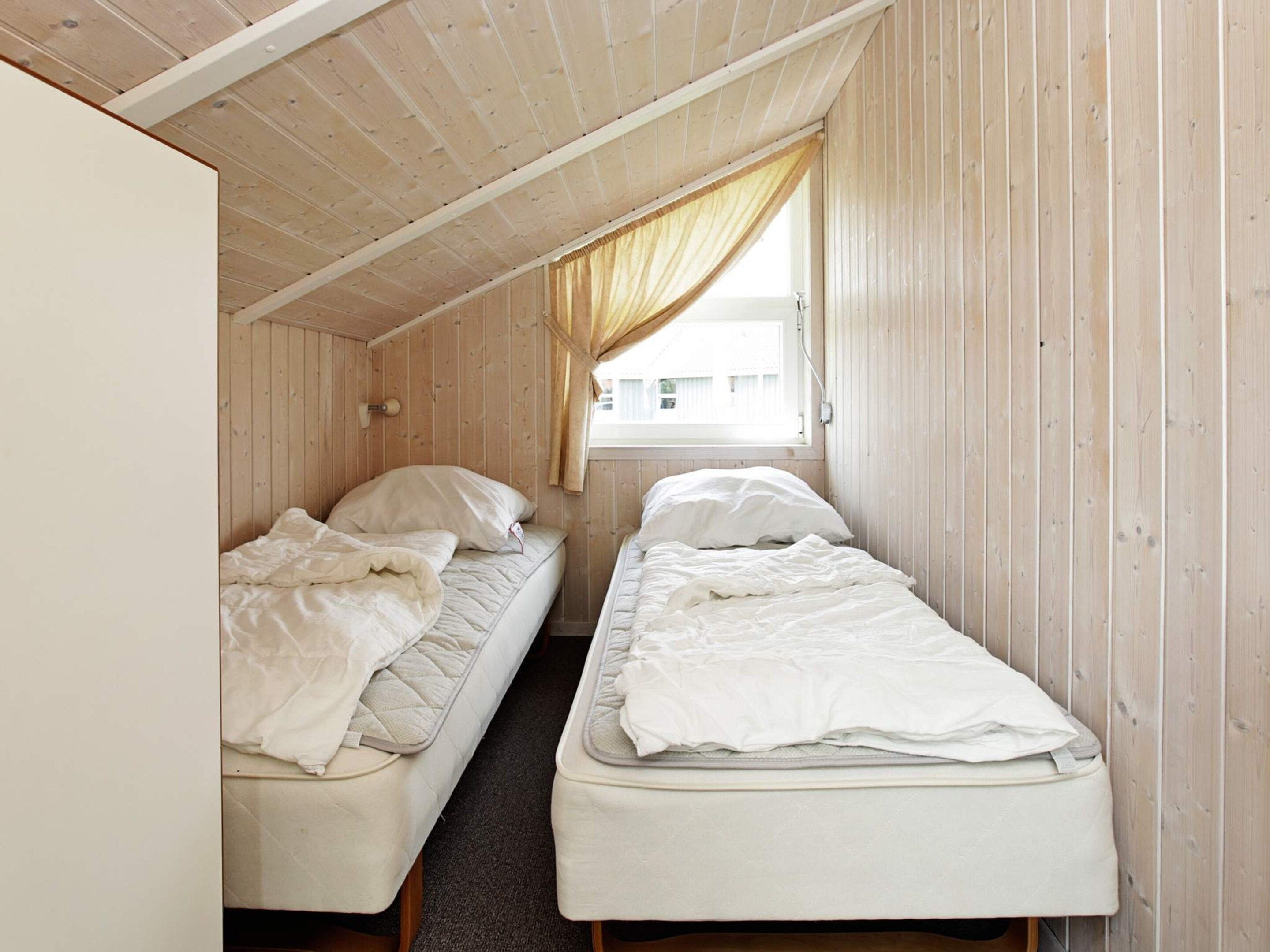 Uma cabana pequena, com um único quarto, duas camas, paredes de madeira e uma janela com cortinas.