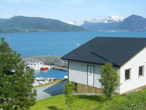 Uma casa com telhado escuro, um cais com barcos e um lago com montanhas ao fundo.