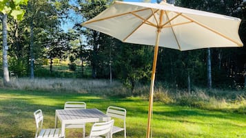 Uma área de refeições ao ar livre com mesa, cadeiras, um grande guarda-sol e um campo gramado com árvores ao fundo.