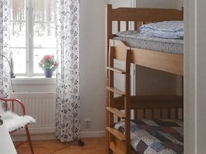 Uma cama beliche com colcha, uma janela com cortinas, um radiador e uma pequena planta.