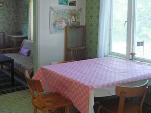 Quarto com uma mesa coberta por um tecido estampado, duas cadeiras de madeira, um sofá com capa estampada, uma cadeira de madeira, uma mesinha e uma janela com uma bandeira.