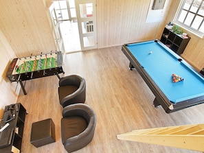 Uma mesa de bilhar com bolas, uma mesa de futebol de mesa e duas poltronas estofadas em um quarto com piso de madeira e uma janela.