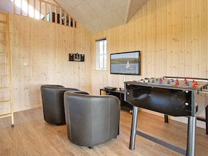 Quarto com paredes de madeira, mesa de futebol, dois pufes pretos, janela com vista para o mar e televisão na parede.