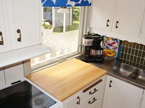 Cozinha compacta com armários brancos, bancada de madeira, cafeteira e janela com vista para uma casa e áreas verdes.