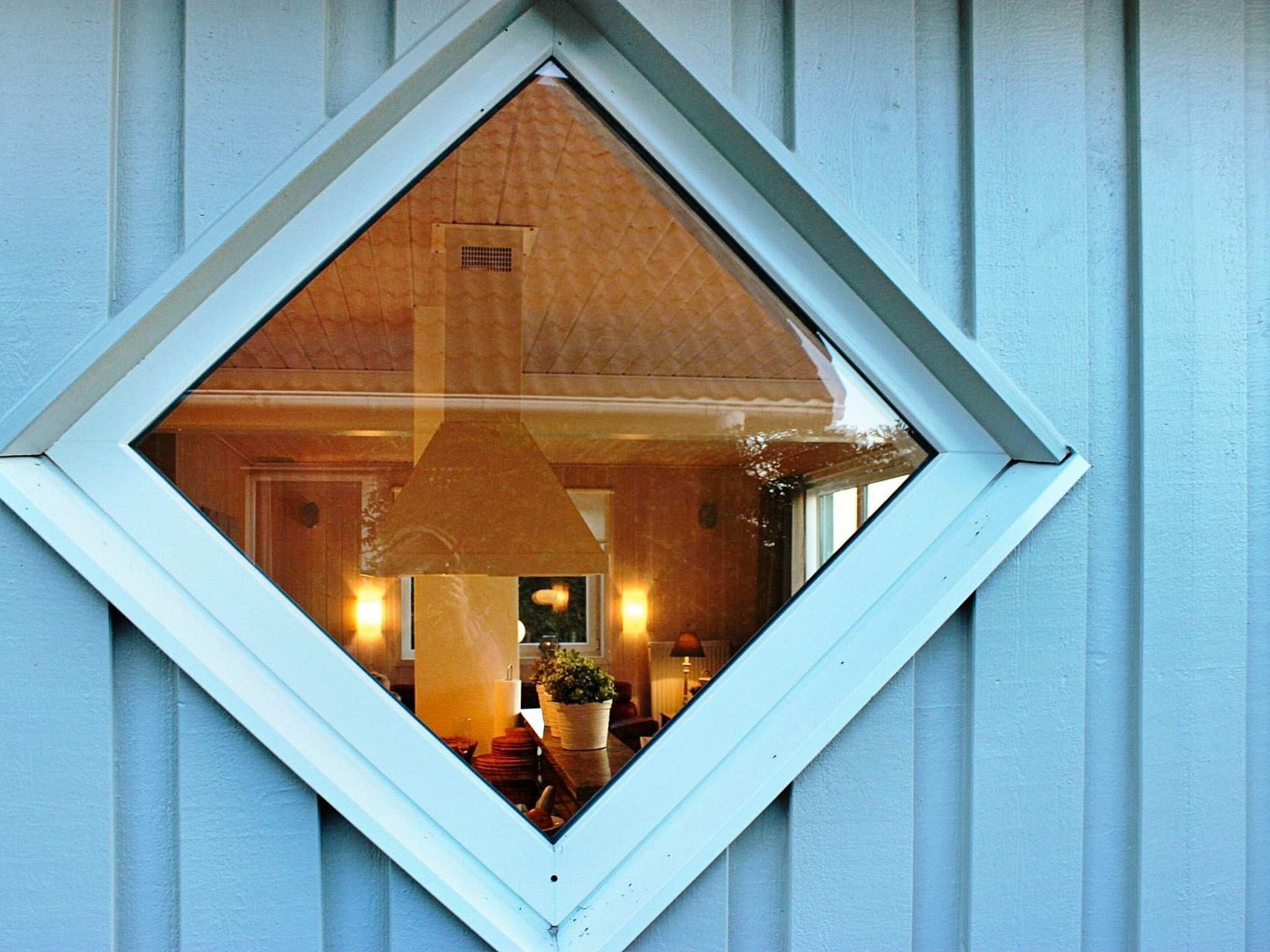 Janela em formato de diamante com vista para um espaço interno, incluindo uma mesa com uma planta e um abajur.