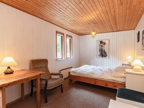 Quarto com teto de madeira, uma cama, uma cadeira, uma mesa e duas janelas.