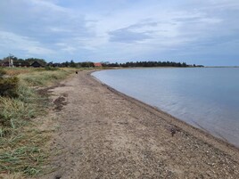 Uma praia arenosa com um lago calmo, algumas casas ao fundo e um céu limpo.
