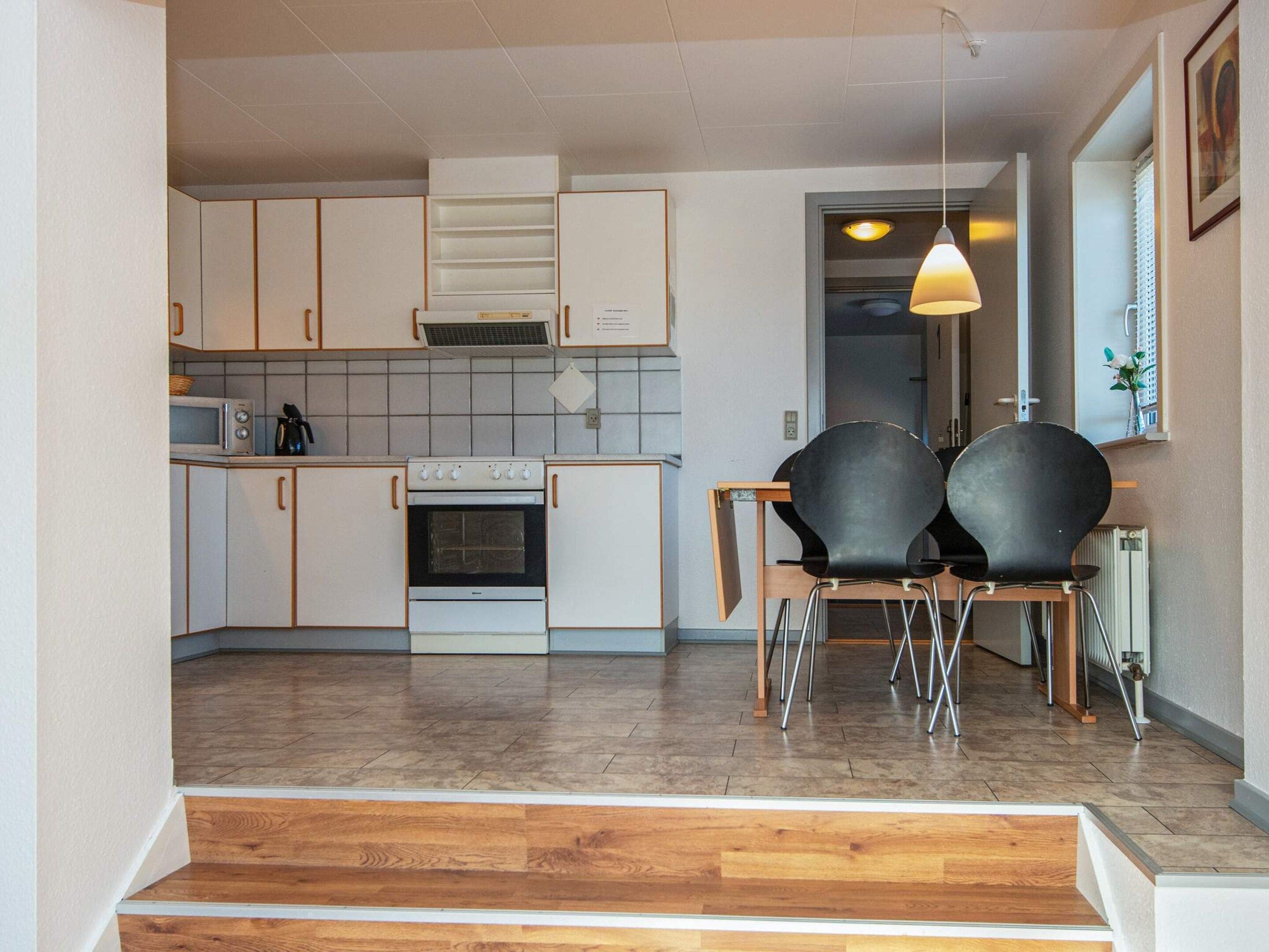 Cozinha moderna com armários brancos, forno embutido e micro-ondas. Possui uma área de jantar com duas cadeiras e uma mesa.
