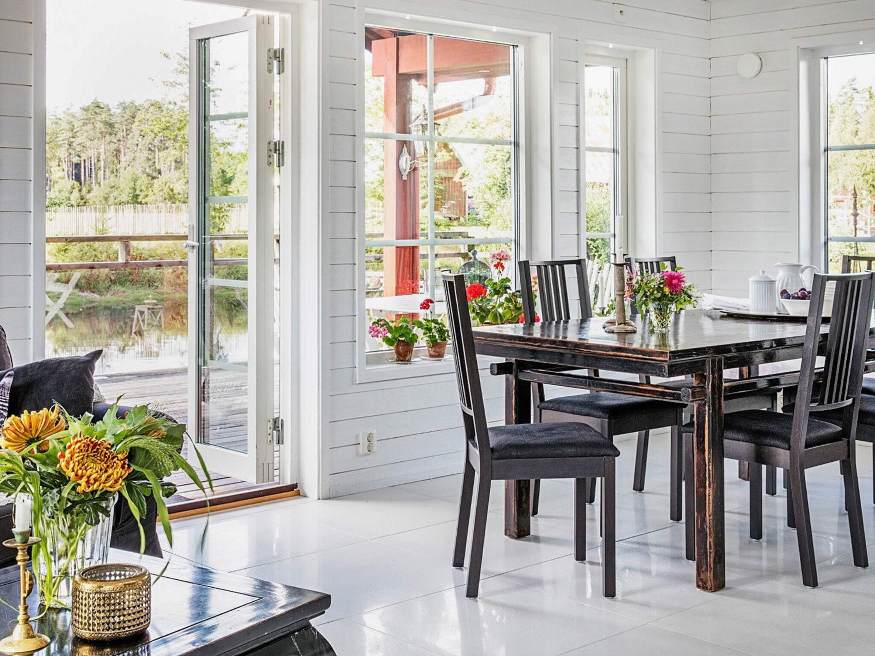 Uma sala de jantar com mesa posta para quatro pessoas, rodeada por cadeiras, e vista para um jardim através de grandes janelas.