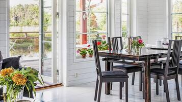 Uma sala de jantar com mesa posta para quatro pessoas, rodeada por cadeiras, e vista para um jardim através de grandes janelas.