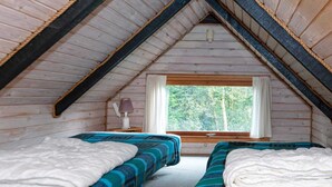Um quarto de sótão aconchegante com duas camas, uma janela com vista para árvores e uma luminária em uma mesa de cabeceira.