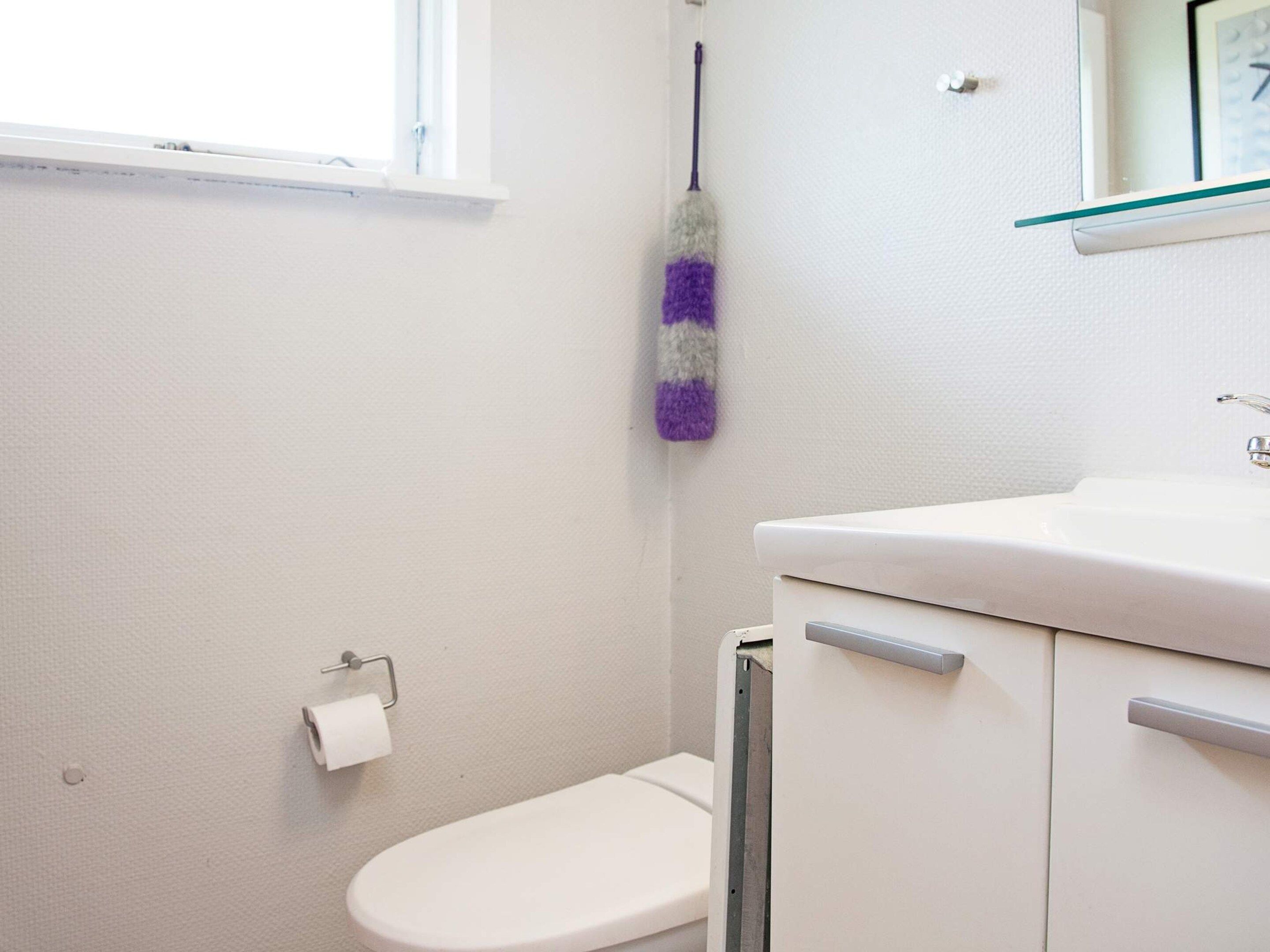 Um banheiro com vaso sanitário, um rolo de papel higiênico, uma prateleira de vidro acima da pia e um esfregão pendurado na parede.