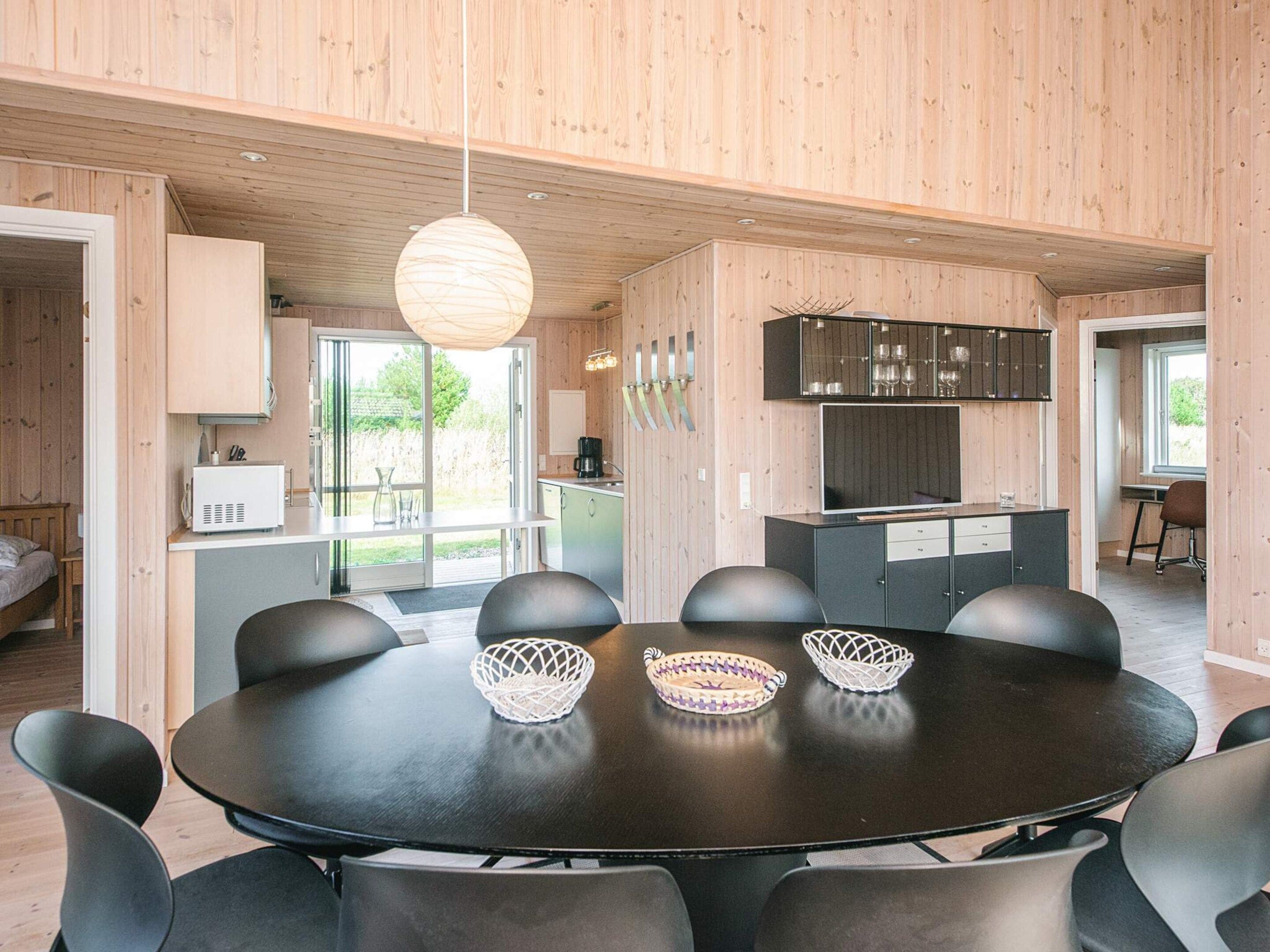 Interior moderno com mesa de jantar redonda, cadeiras pretas, área de cozinha com armários e luminária pendente.