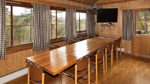 Quarto com painéis de madeira, uma mesa comprida com cadeiras, televisão de tela plana na parede e janelas amplas com cortinas xadrez.