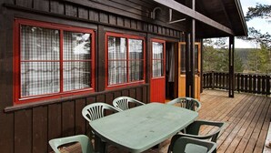 Uma cabana de madeira com varanda, uma mesa com cadeiras e grandes janelas com molduras vermelhas.