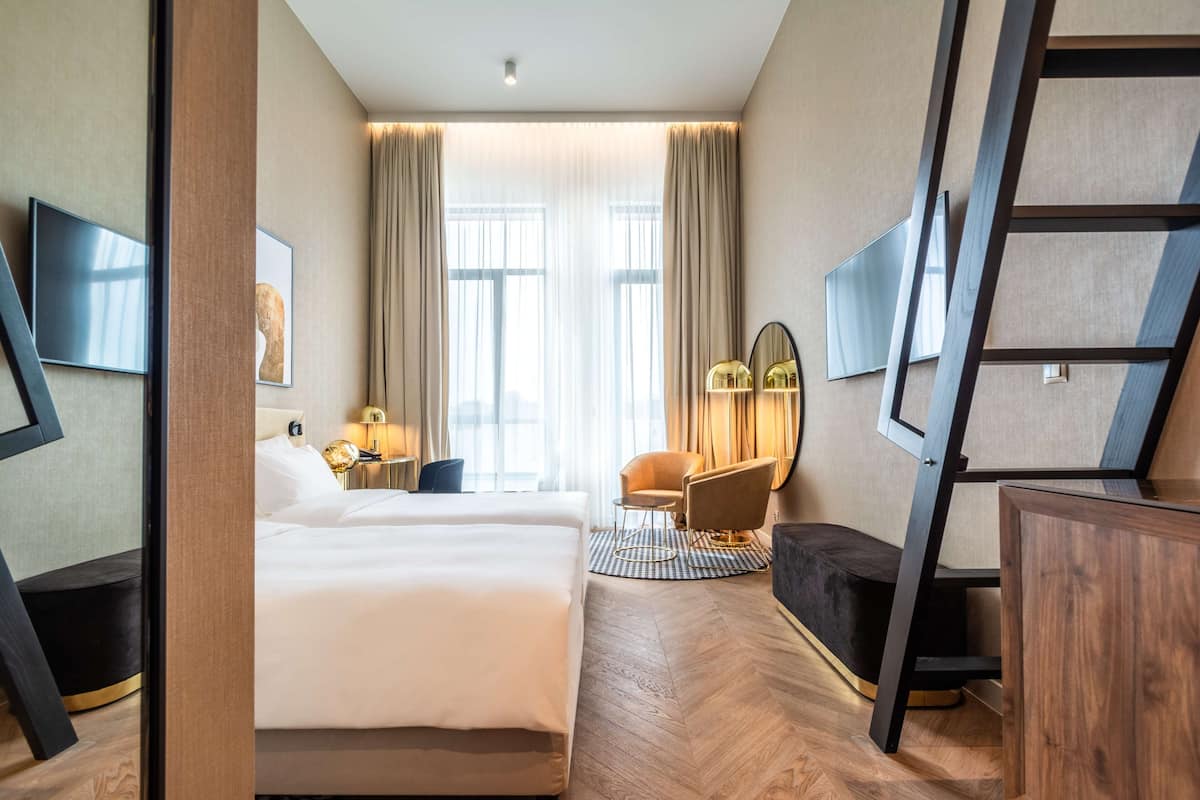 Quarto de hotel com uma cama grande, uma escrivaninha com cadeira, televisão na parede e uma janela com cortinas.