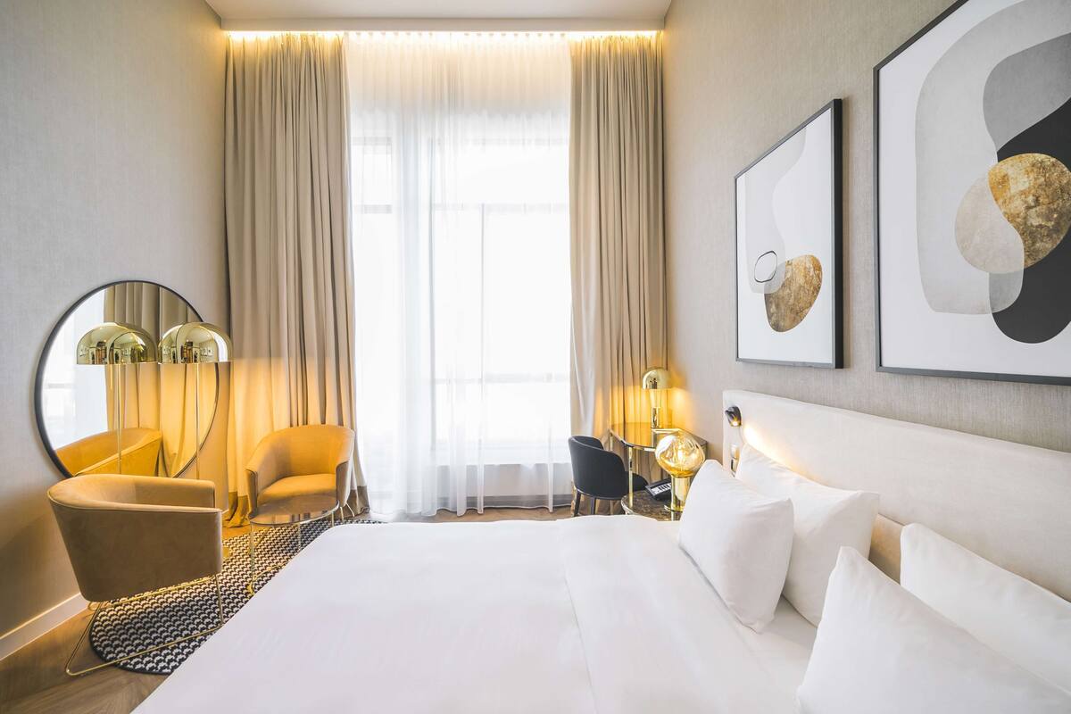 Quarto de hotel com uma cama grande, duas poltronas, uma escrivaninha e uma janela ampla com cortinas.