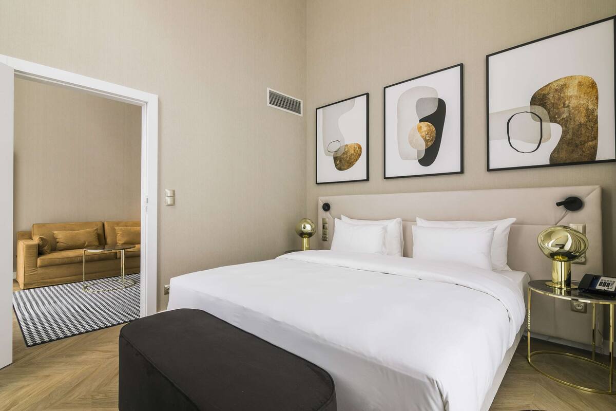 Quarto de hotel com uma cama grande, mesa de cabeceira com abajur, uma mesinha e um sofá na sala adjacente.
