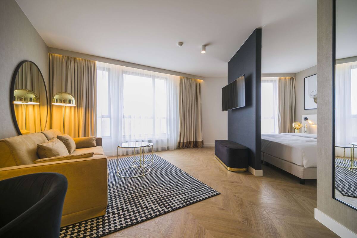 Quarto de hotel moderno com sofá, cama, televisão fixada na parede e uma janela grande com cortinas.