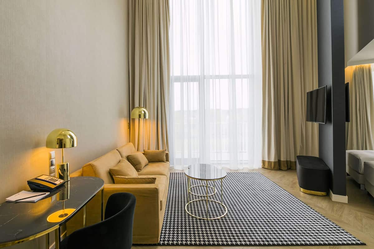 Quarto de hotel moderno com sofá, mesinha redonda, escrivaninha com abajur, televisão e tapete estampado.