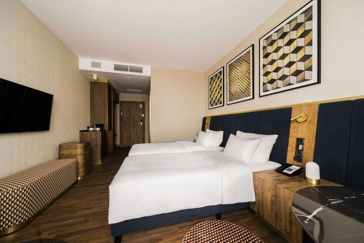 Quarto de hotel com uma cama grande, uma mesinha de cabeceira com abajur, uma televisão de tela plana na parede e um banco de madeira.
