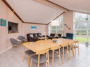 Uma sala de estar moderna com piso de madeira, um sofá de couro marrom, uma poltrona azul, uma mesa de jantar com oito cadeiras e grandes janelas com vista para o exterior.