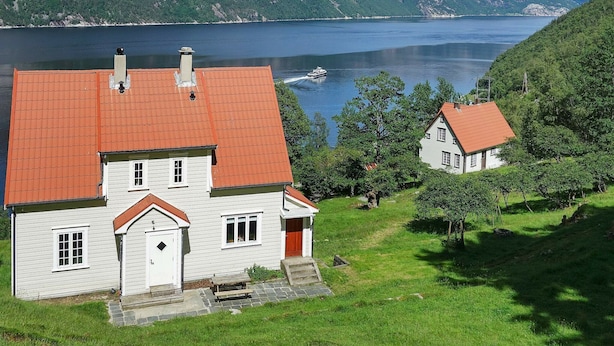 Um edifício branco com telhado vermelho, localizado numa encosta gramada com vista para um lago e montanhas ao fundo.
