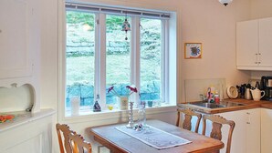 Uma área de refeições com uma mesa posta para quatro pessoas, uma janela com vista para o verde e uma cozinha com armários e eletrodomésticos.