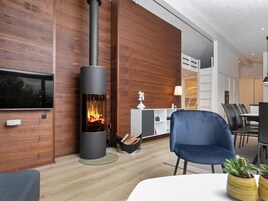 Uma sala de estar moderna com uma parede revestida de madeira, uma lareira cilíndrica preta, uma televisão de tela plana, uma poltrona azul e uma área de jantar com mesa e cadeiras.