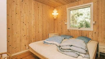 Quarto com teto de madeira, cama com almofadas, mesa de cabeceira e janela com vista para árvores.