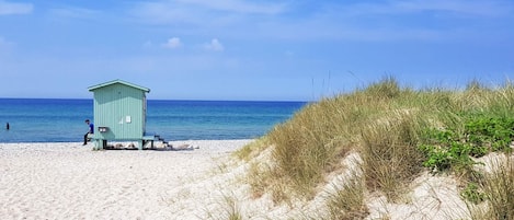 Uma praia com uma pequena estrutura azul, possivelmente um posto de salva-vidas, e uma pessoa parada perto.