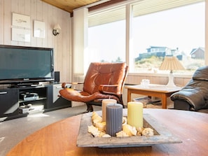 Uma sala de estar com televisão, poltronas de couro, uma mesa de centro com velas e uma janela com vista para casas.