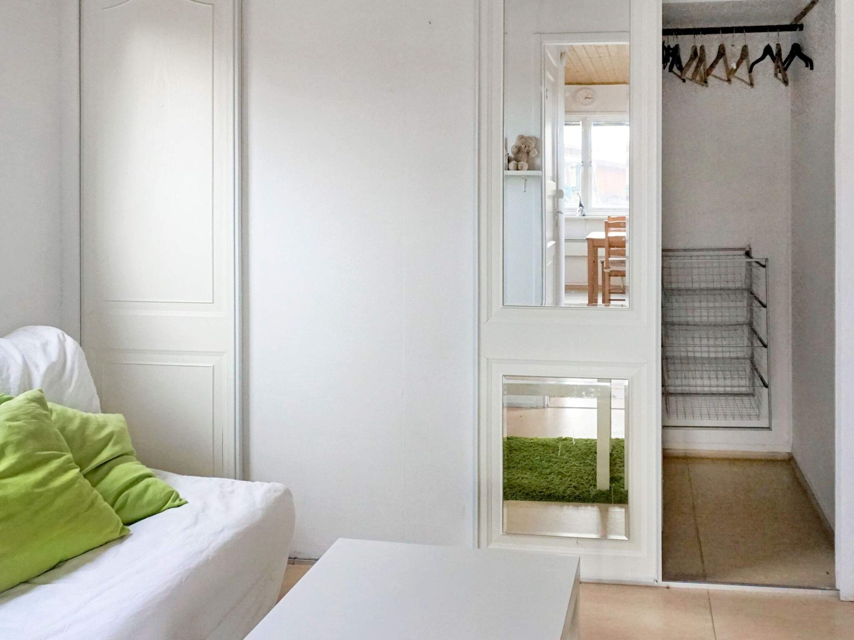 Um quarto com cama, mesa de cabeceira branca, travesseiro verde, parede branca, porta e vista para uma cozinha e um lance de escadas.