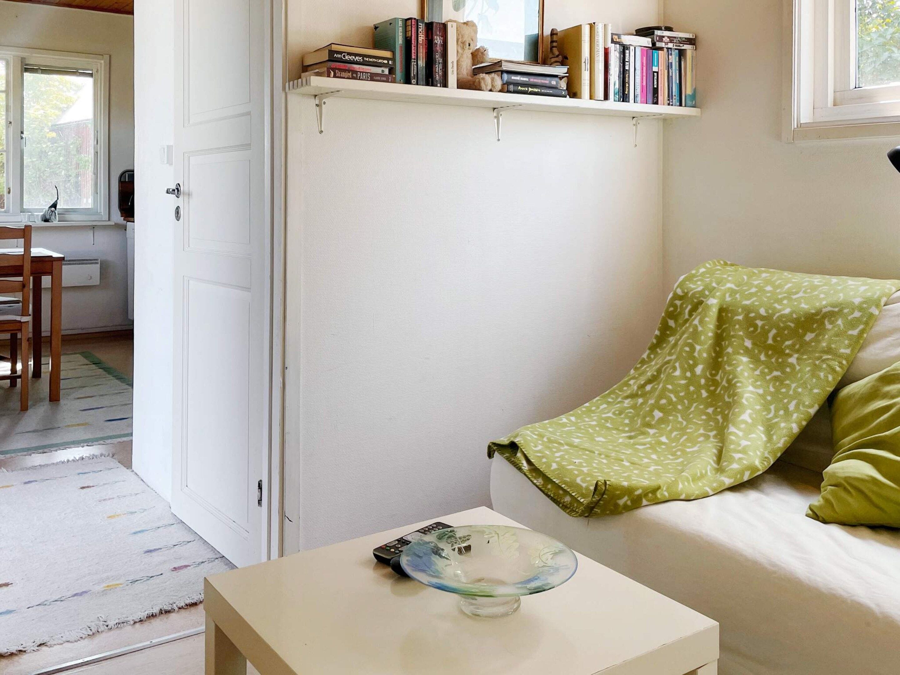Quarto com cama, mesa de cabeceira com tigela e controle remoto, estante e janela com vista para árvores.