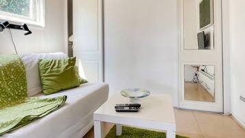 Quarto minimalista com sofá branco, uma mesinha branca com uma tigela e controles remotos, e um tapete verde.