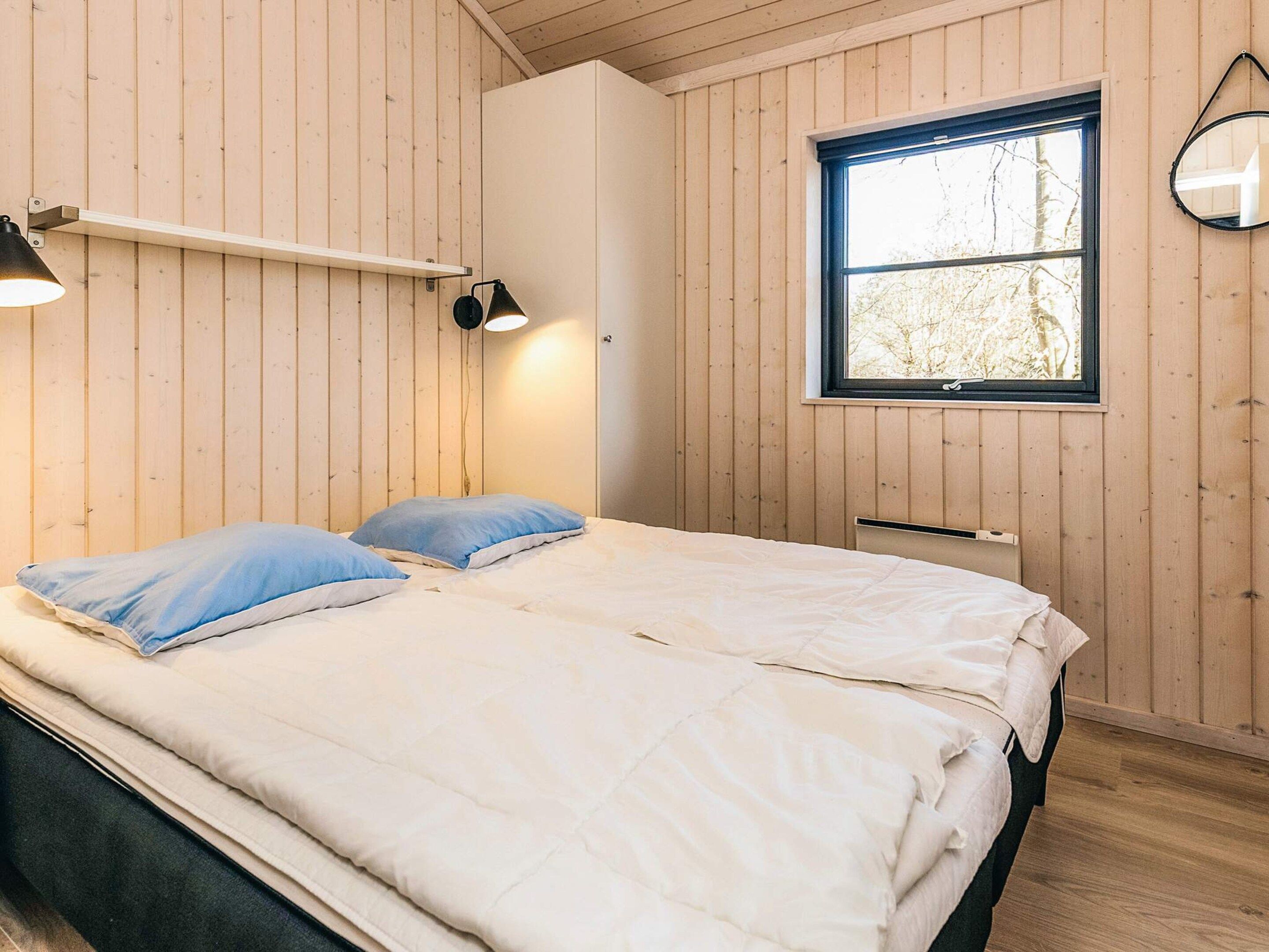 Quarto com cama, duas almofadas, mesa de cabeceira, abajur, janela e paredes de madeira.