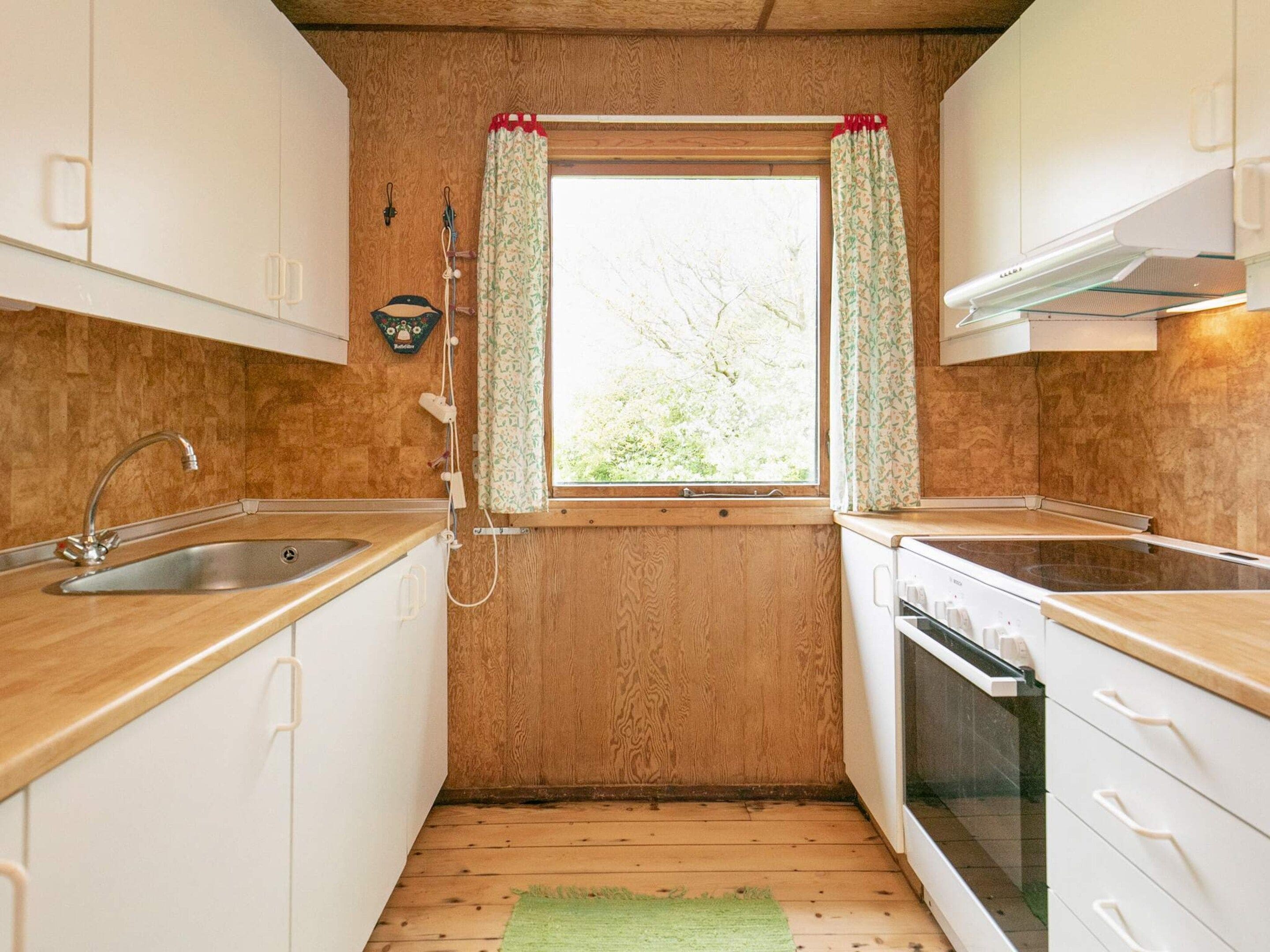 Cozinha compacta com armários de madeira, pia, janela com cortinas e fogão.