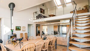 Uma ampla área de refeições com uma longa mesa de madeira e cadeiras, uma cozinha com eletrodomésticos modernos e uma escada em espiral que leva a um andar superior.