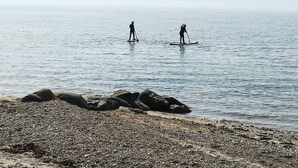 Duas pessoas praticam paddleboard em águas calmas, com uma margem rochosa em primeiro plano.