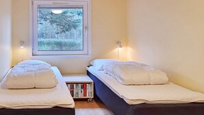 Um quarto pequeno com duas camas, uma janela com vista para árvores e uma estante com livros.