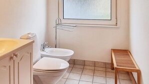 Casa de banho pequena com sanita, bidé e lavatório. Há uma janela sobre o bidé e um banco de madeira à direita.